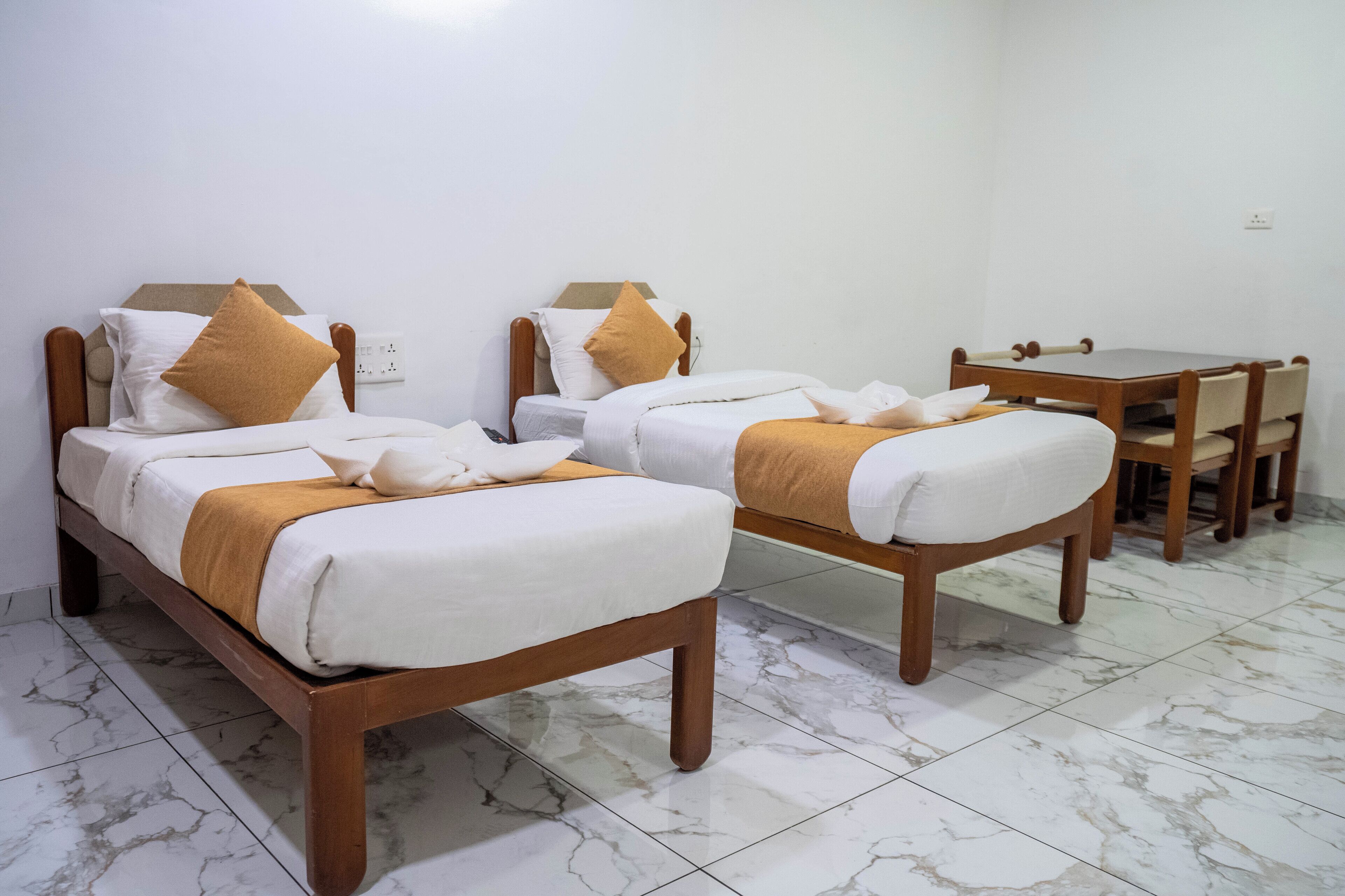 Quarto com duas camas de madeira, cada uma com roupa de cama branca e um cobertor marrom. Há uma mesa de madeira e cadeiras no quarto.