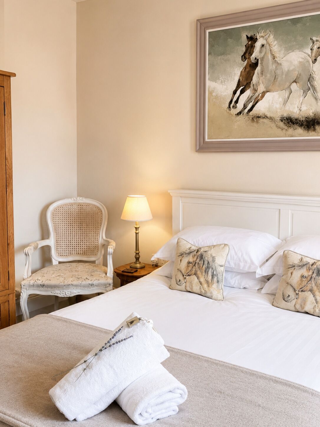Quarto de hotel com cama, uma cadeira, um abajur e uma pintura de cavalos na parede.