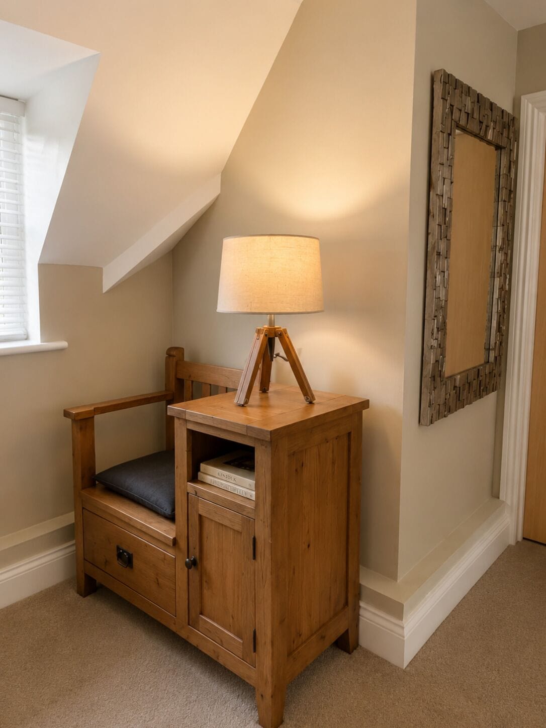 Quarto nos cantos com teto inclinado, uma cadeira de madeira com espaço de armazenamento, um abajur e um espelho na parede.