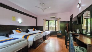 Quarto de hotel com três camas, um ventilador de teto, ar condicionado, uma mesa pequena com uma chaleira e um espelho grande.