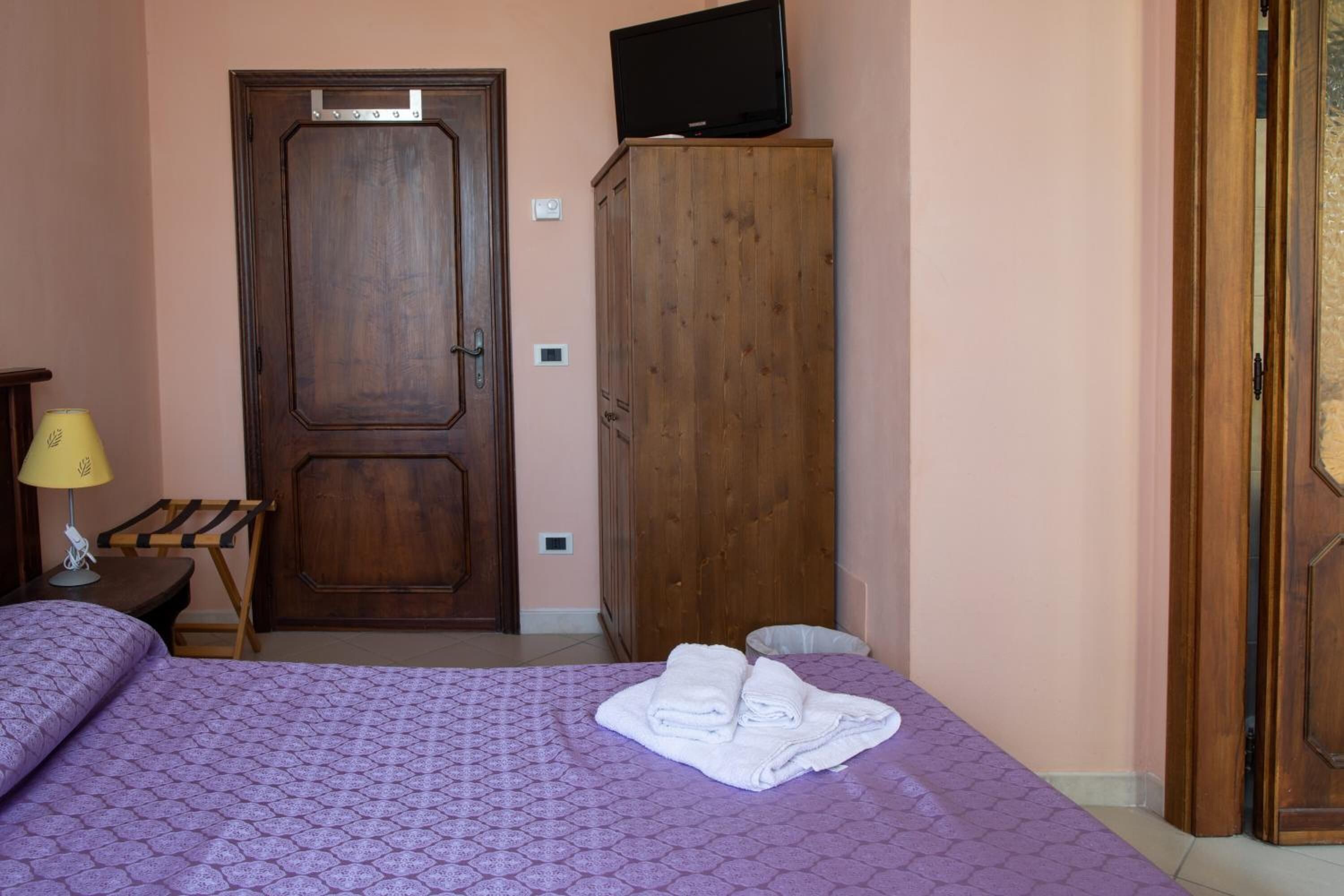 Quarto de hotel com uma cama, um guarda-roupa de madeira, uma televisão em cima do guarda-roupa e um abajur sobre uma mesinha de cabeceira.