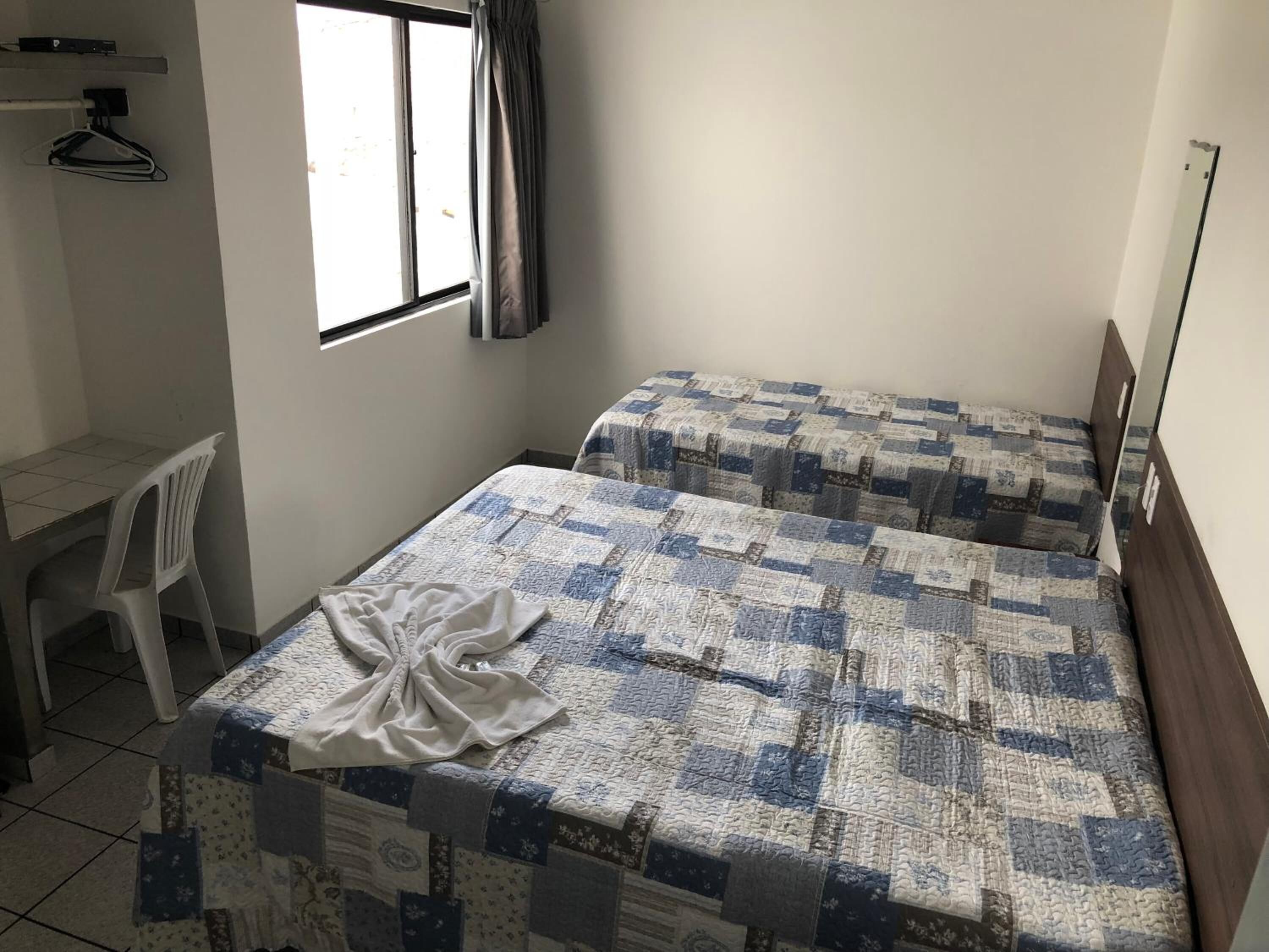 Quarto pequeno e limpo, com duas camas, uma escrivaninha e uma cadeira.