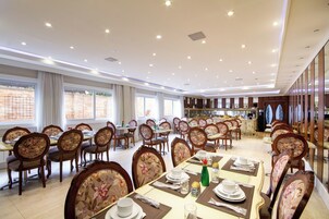 Uma sala de jantar ampla, com várias mesas e cadeiras, janelas grandes e uma cozinha ao fundo.
