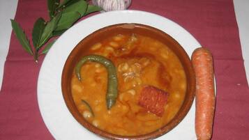 Uma tigela de sopa com legumes e uma salsicha, colocada sobre um prato branco com um fundo de toalha vermelha.