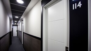 Um corredor moderno de hotel com piso de madeira escura, paredes brancas e uma porta preta com a placa '114'.