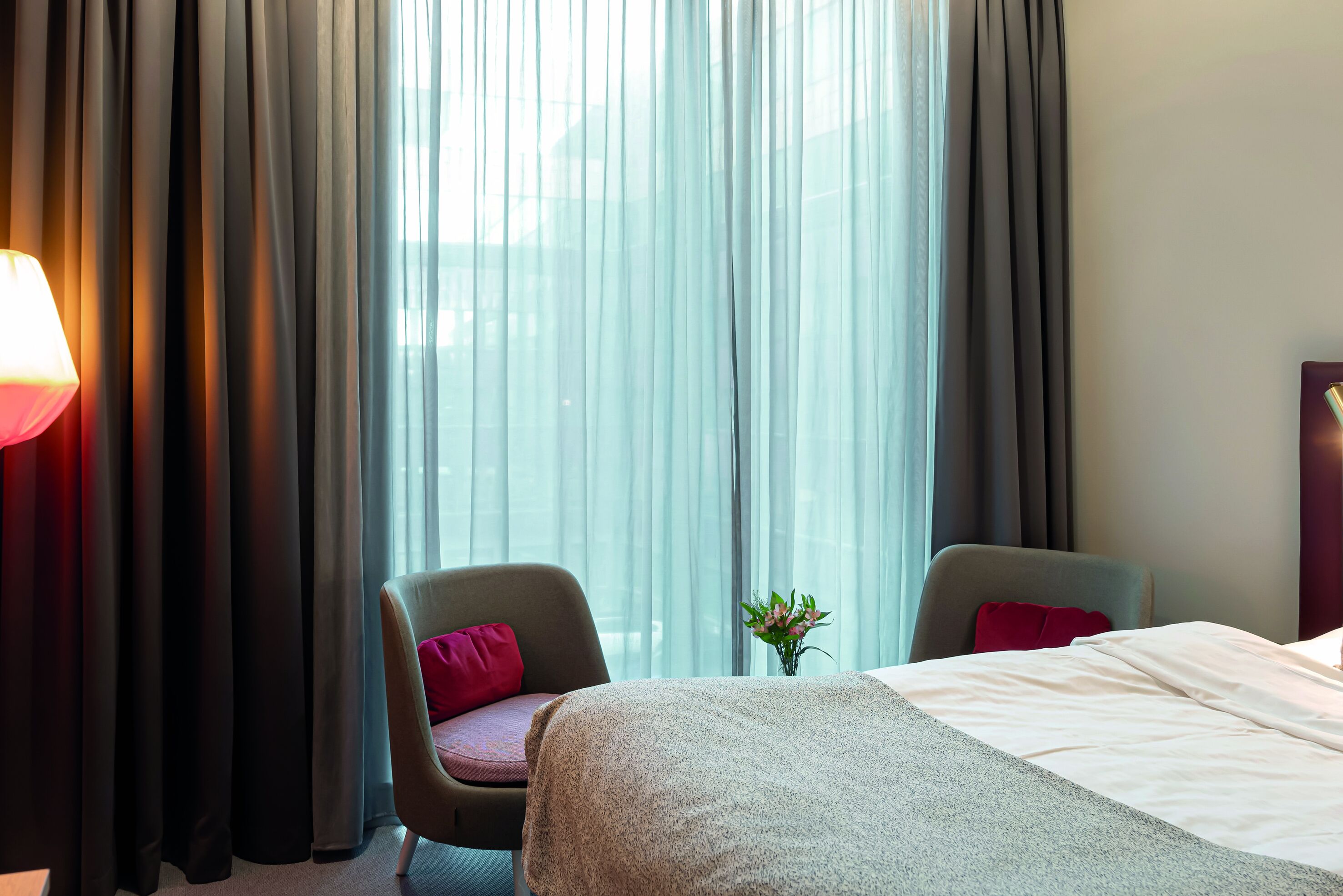 Quarto de hotel com cama, duas cadeiras, abajur e janela com cortinas.
