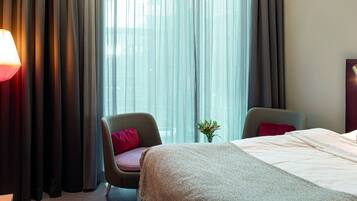 Quarto de hotel com cama, duas cadeiras, abajur e janela com cortinas.