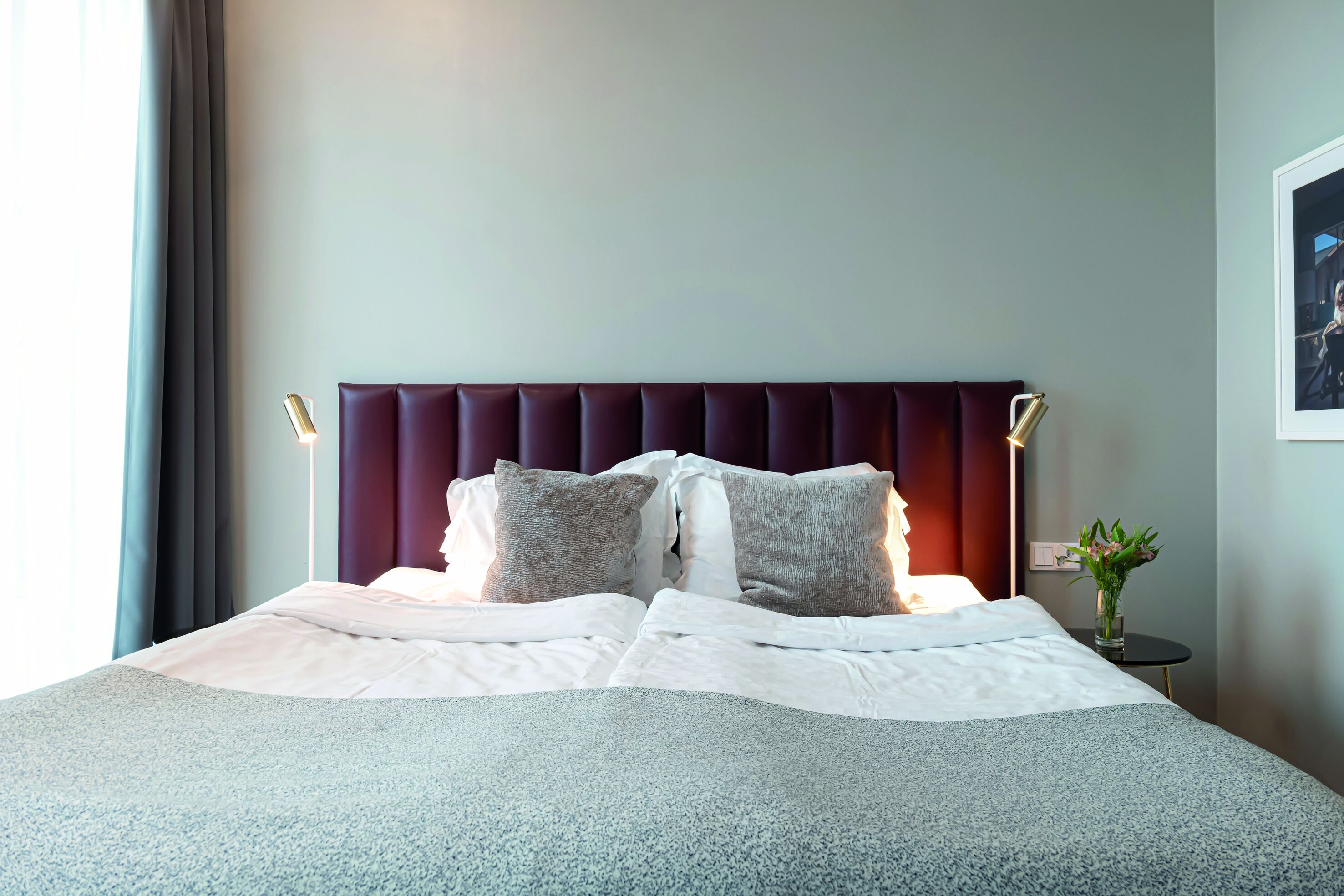 Uma cama bem arrumada com lençóis brancos e um cobertor cinzento, uma cabeceira escura e uma pequena mesa de cabeceira com um vaso de flores.