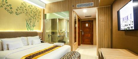 Quarto de hotel com uma cama grande, um banco de madeira, uma televisão e um box de chuveiro em vidro.