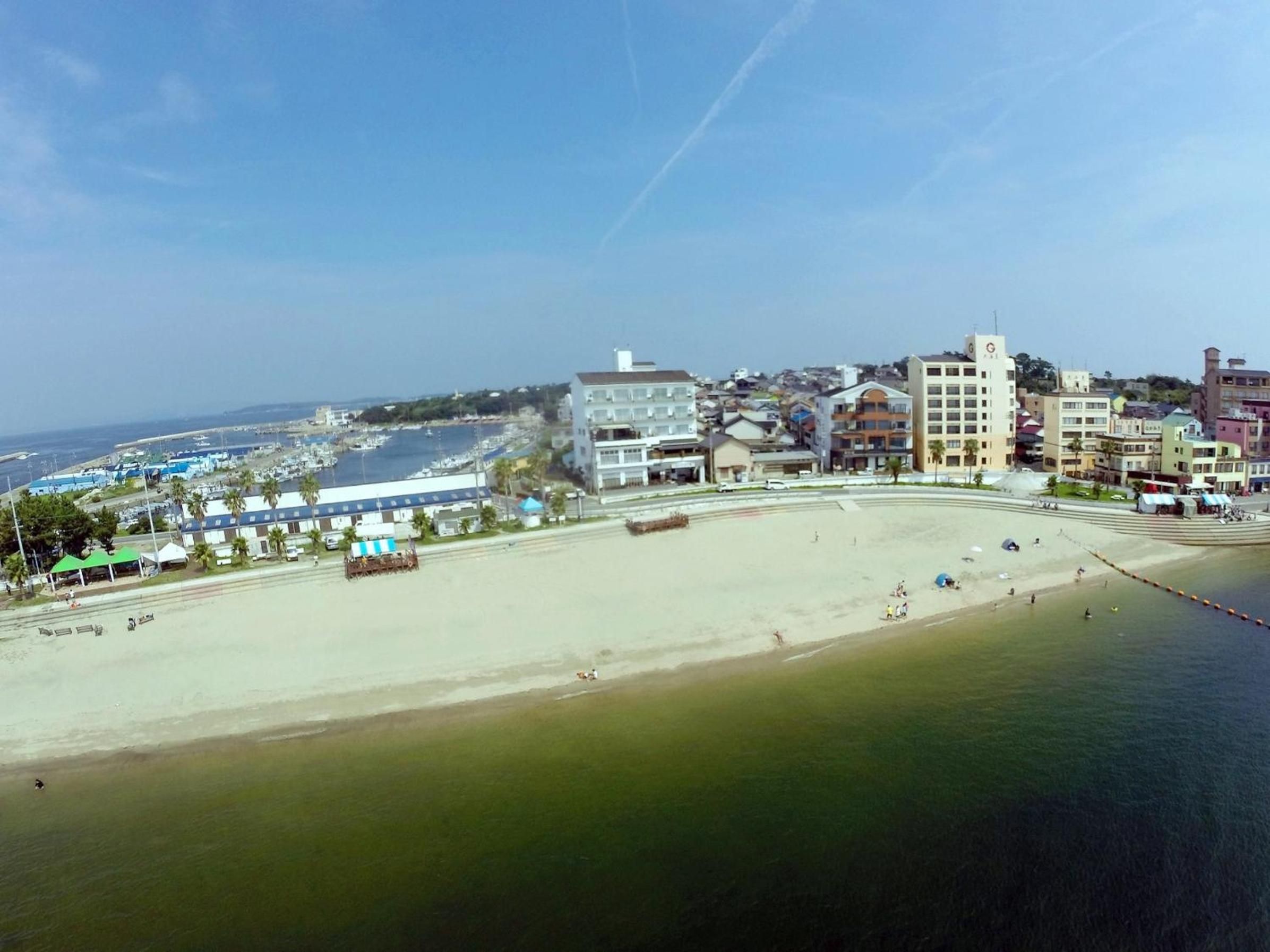 Paisagem costeira com uma extensa praia arenosa, um porto de iates e diversos edifícios, incluindo um hotel imponente com uma torre.