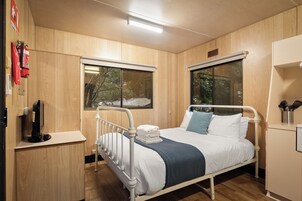 Quarto individual com uma cama, chão de madeira, mesa de cabeceira, janela e vista para árvores.