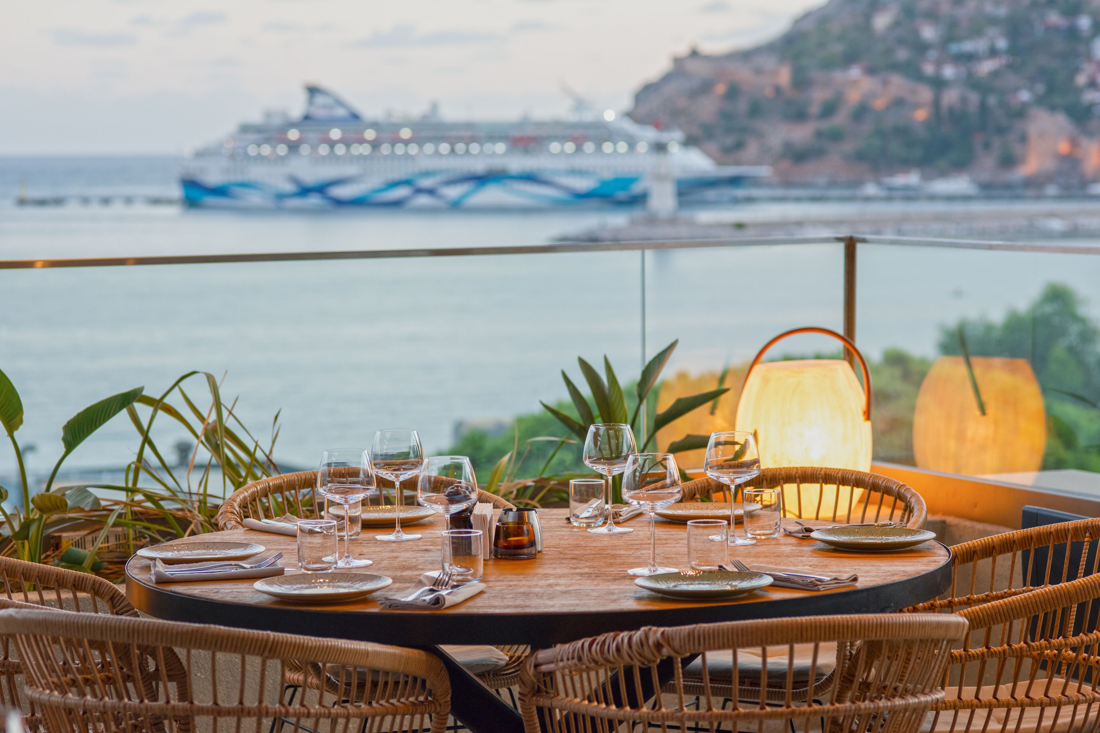 Uma mesa de jantar posta com pratos, copos e talheres, com vista para um corpo de água onde se avista um cruzeiro ao longe.