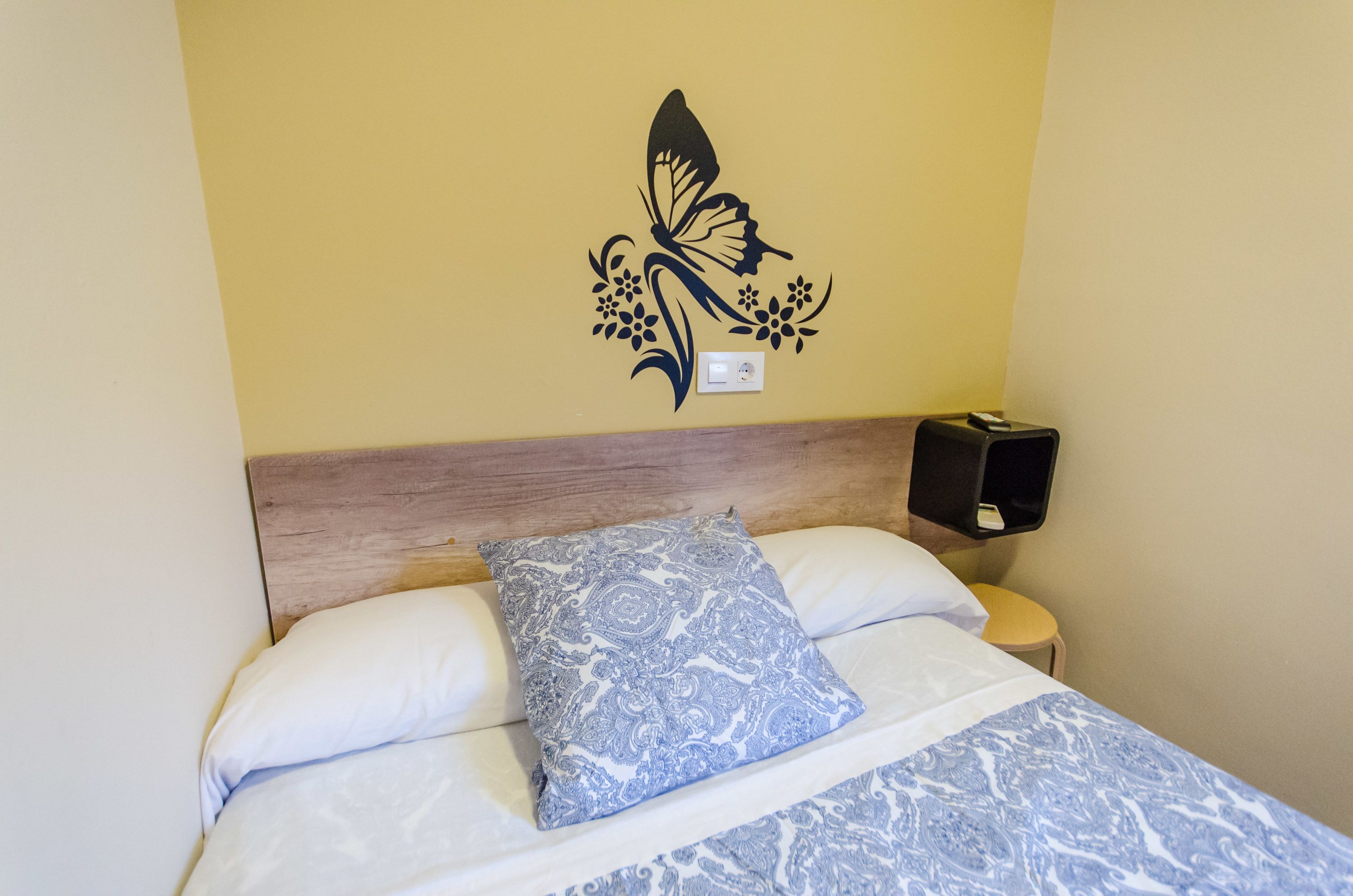 Uma cama com travesseiro estampado, um alto-falante fixado na parede e um adesivo decorativo de borboleta.