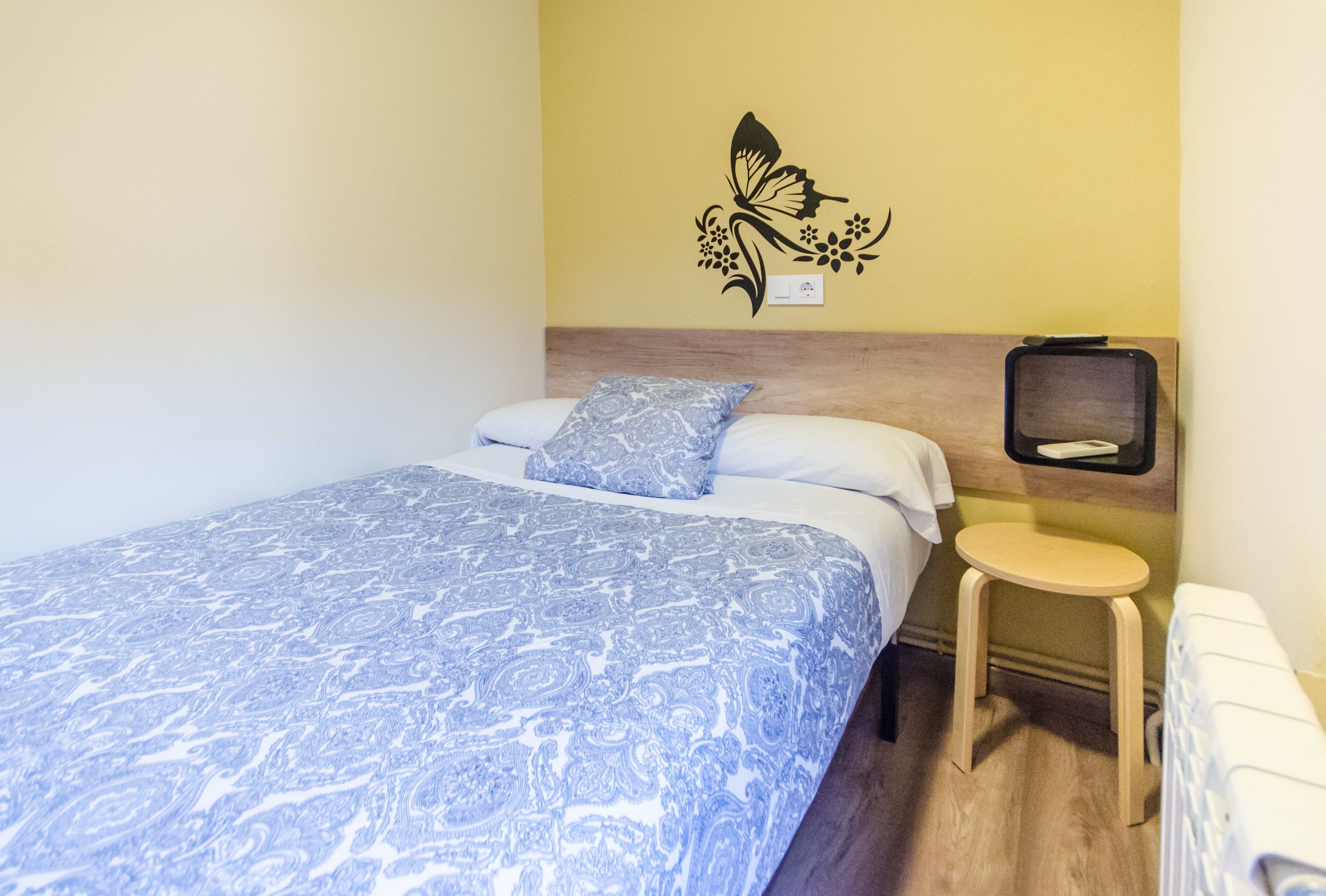 Cama bem arrumada com colcha azul estampada, almofada combinando e mesa de cabeceira fixa na parede com um pequeno banquinho.