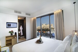 Quarto de hotel com uma cama grande, uma escrivaninha com computador, uma pequena planta e vista para um edifício histórico.