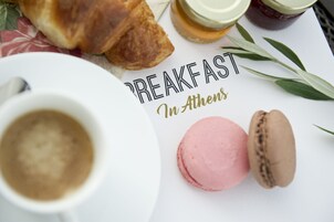 Uma mesa de café da manhã com um croissant, dois macarons e uma xícara de café, sobre um jogo americano com a inscrição “BREAKFAST In Athens”.