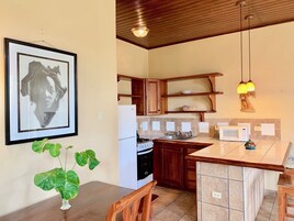 Uma cozinha com armários de madeira, uma geladeira branca, uma pia e um micro-ondas. Há uma planta no balcão e um lustre.