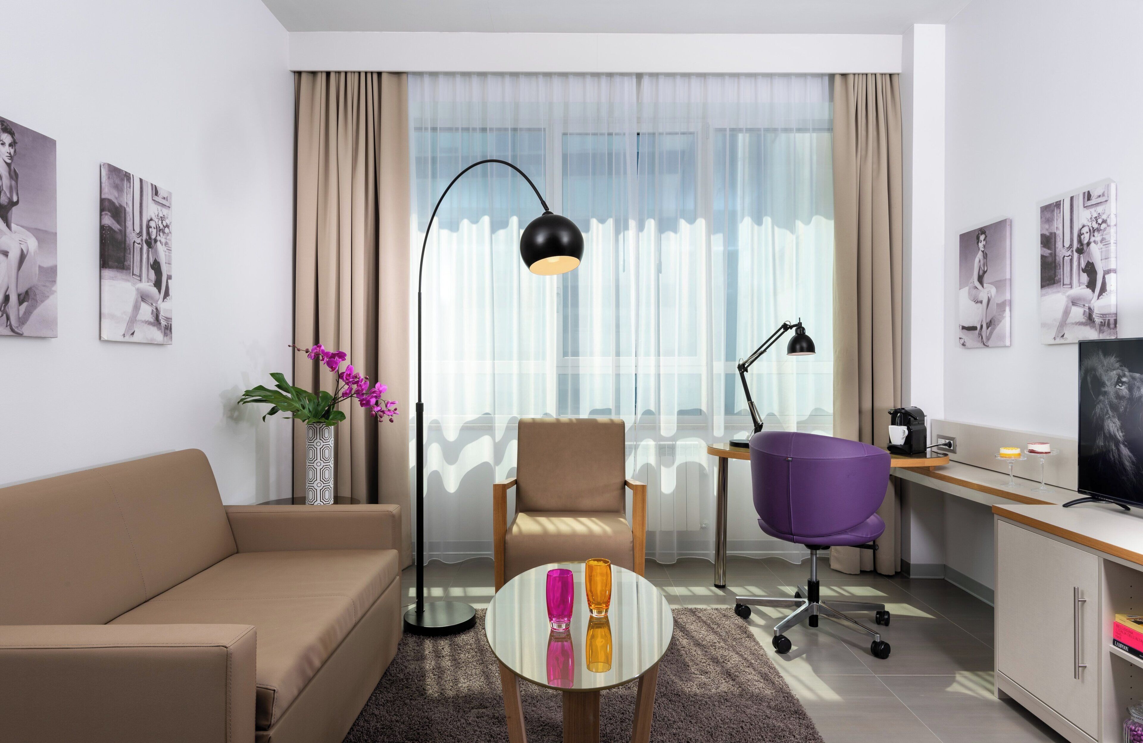 Quarto de hotel moderno com um sofá bege, uma mesa de vidro redonda com dois vasos, uma cadeira de escritório roxa, uma escrivaninha com um computador e uma janela grande com cortinas translúcidas.