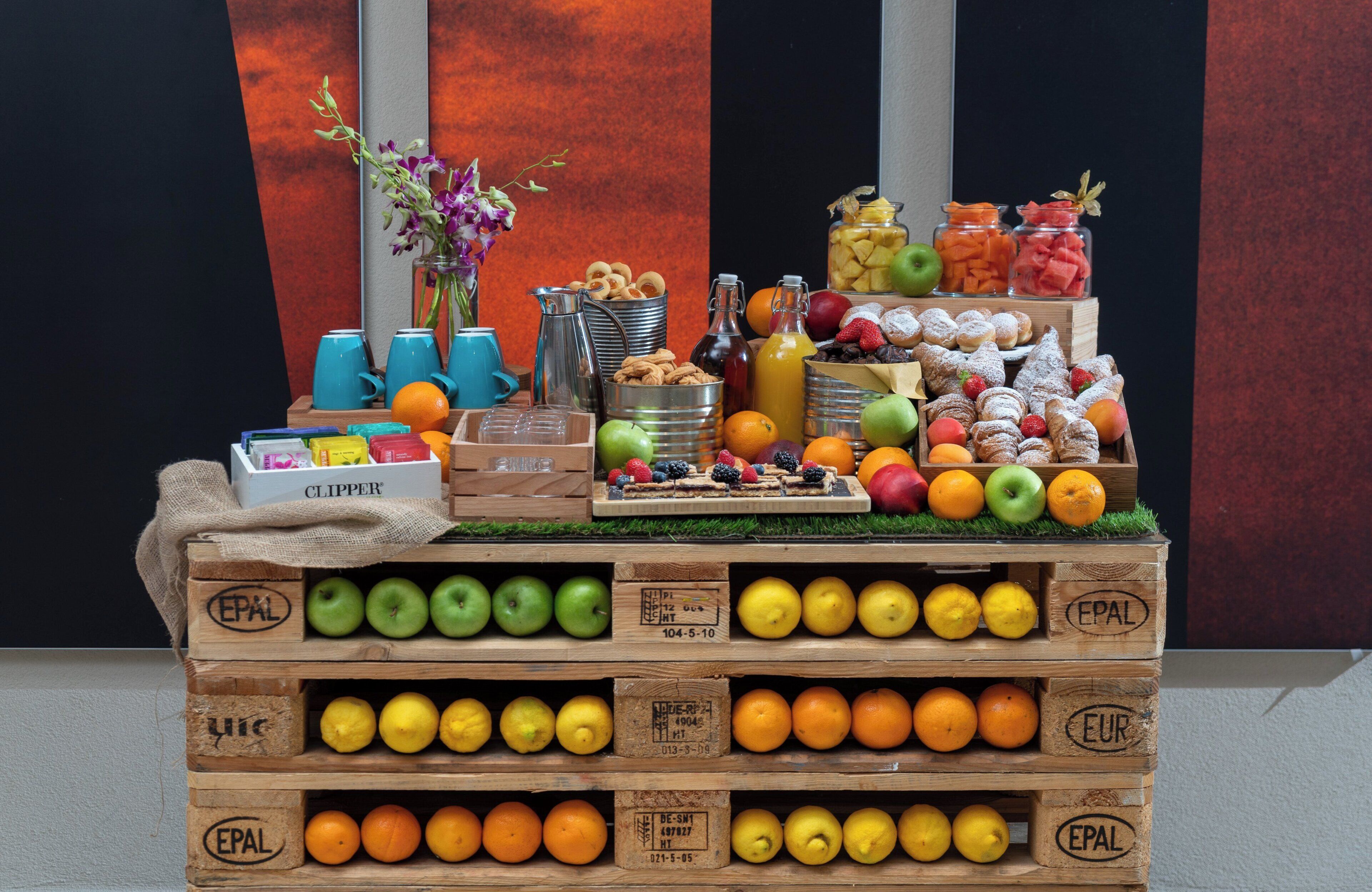 Exposição em caixa de madeira com frutas e lanches, incluindo maçãs, laranjas e diversas frutas em conserva, com um vaso de flores e uma cafeteira ao fundo.