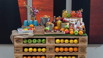 Exposição em caixa de madeira com frutas e lanches, incluindo maçãs, laranjas e diversas frutas em conserva, com um vaso de flores e uma cafeteira ao fundo.
