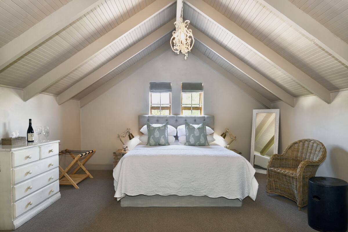 Quarto com teto inclinado, cama com colcha branca e duas almofadas, cômoda, cadeira de vime e um espelho.