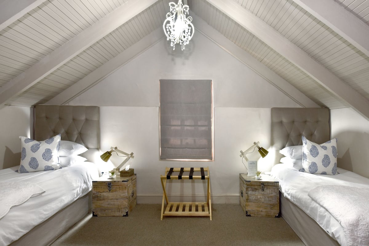 Quarto com duas camas, uma mesinha de cabeceira de madeira, luminária pendente e quadro na parede.