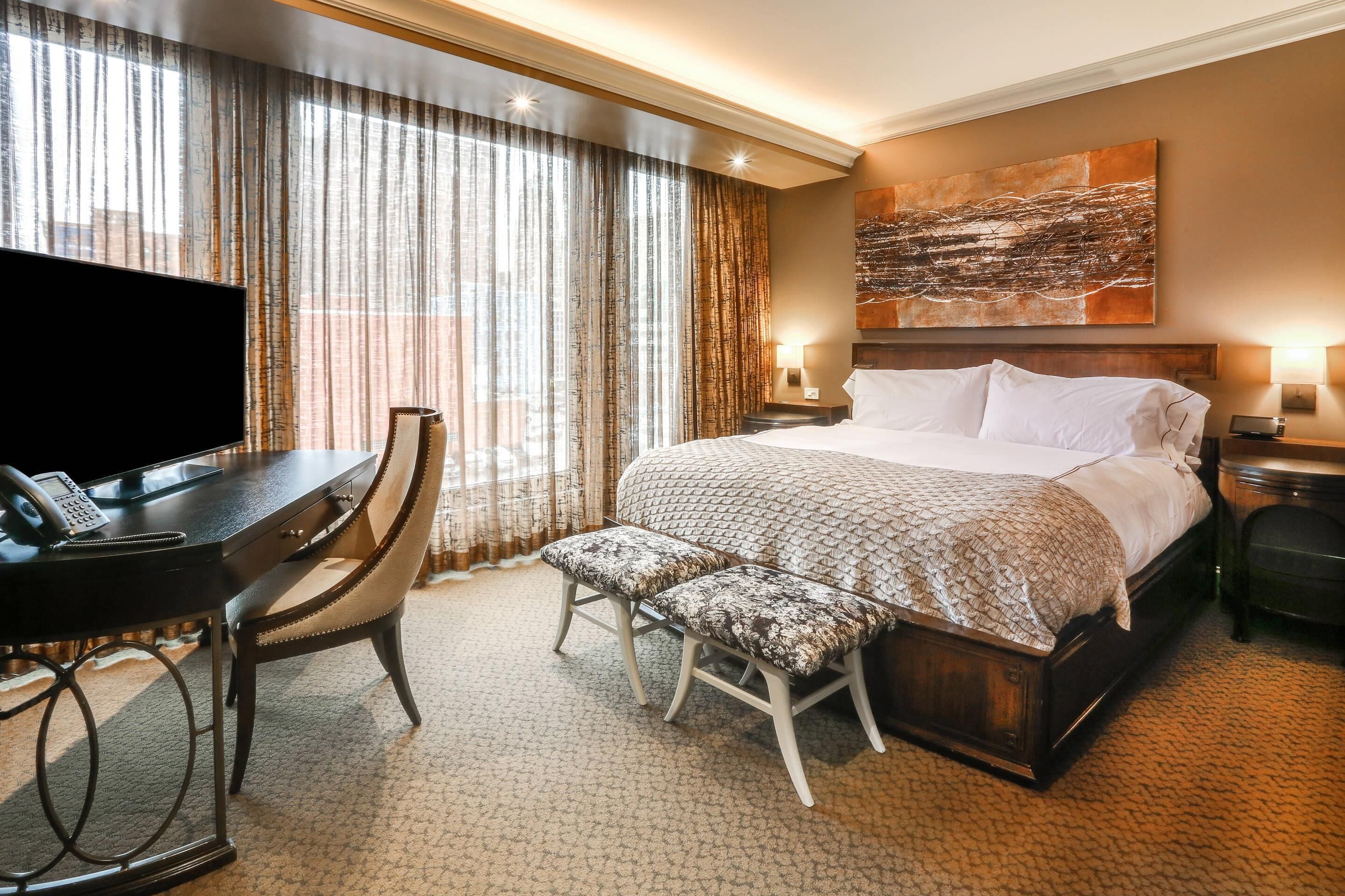 Quarto de hotel com cama, escrivaninha com telefone, uma cadeira, um banco e uma grande pintura abstrata na parede.