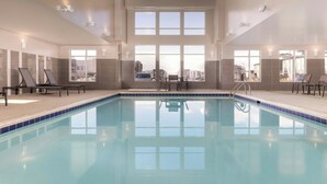 Piscina interna com janelas amplas, cadeiras de estar e vista para a paisagem urbana.