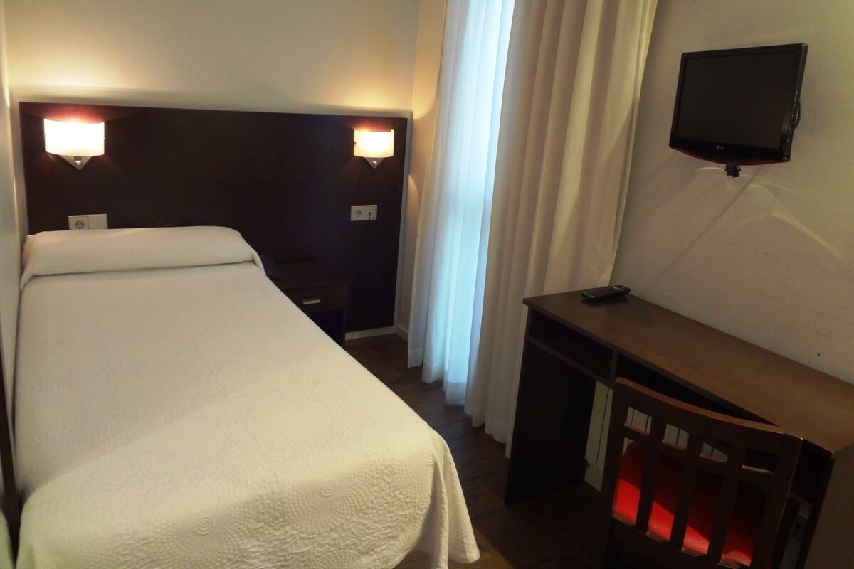 Quarto de hotel com cama, luminárias laterais, escrivaninha com TV e cadeira com iluminação vermelha.
