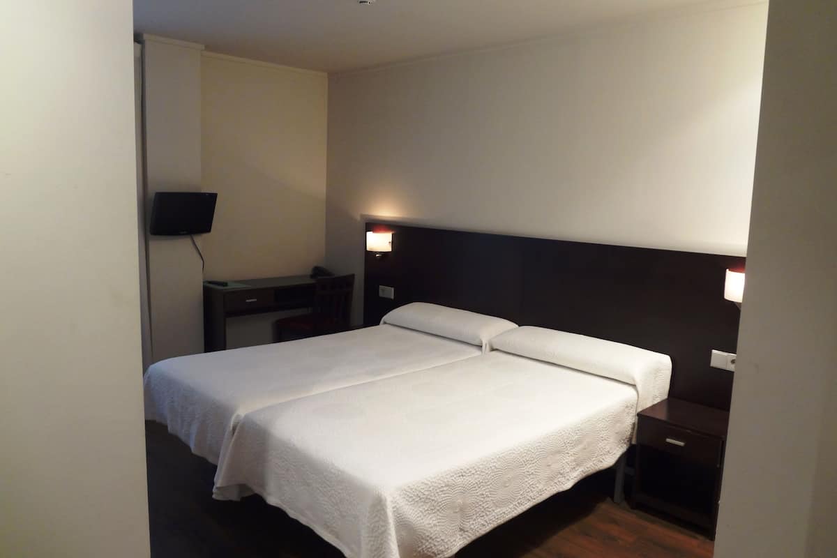 Quarto de hotel com cama, mesas de cabeceira, escrivaninha e televisão na parede.
