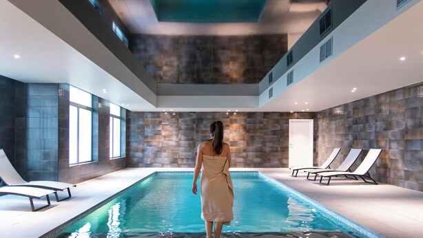 Piscina interior moderna com uma mulher na beira, espreguiçadeiras e uma parede de pedra.
