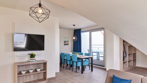 Uma sala de estar moderna com televisão de tela plana, sofá, mesa de centro com revistas e um espaço de jantar com cadeiras azuis e uma mesa.