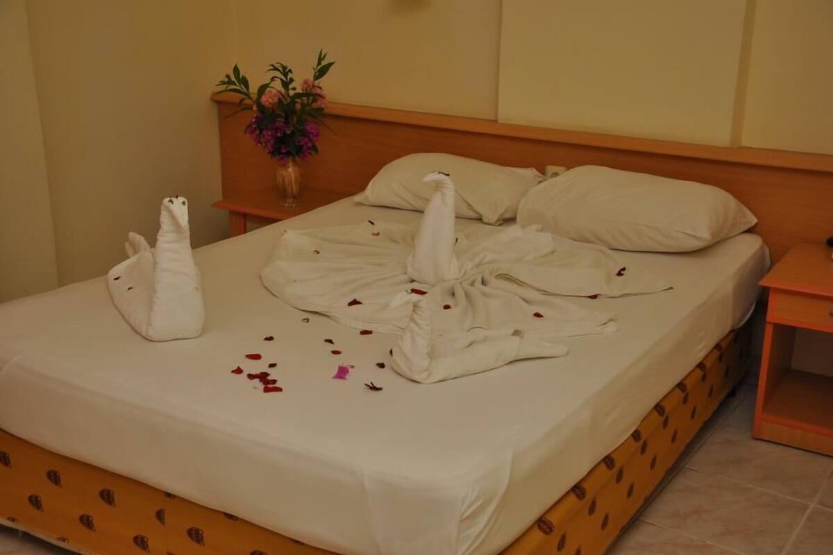 Uma cama com toalhas brancas dispostas em forma de cisnes, um vaso com flores e uma cabeceira de madeira.