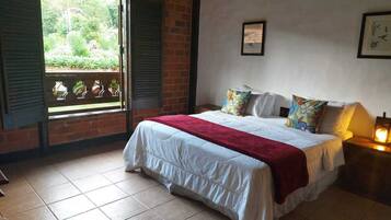 Um quarto com uma cama, almofadas, mesa de cabeceira com abajur, janela com persianas e um quadro na parede.