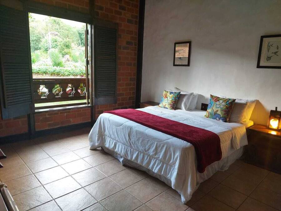 Um quarto com uma cama, almofadas, mesa de cabeceira com abajur, janela com persianas e um quadro na parede.