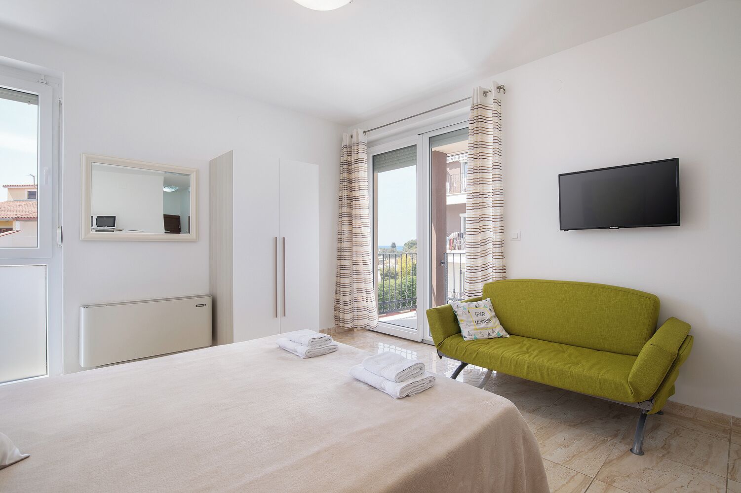 Quarto de hotel com cama, sofá verde, televisão de tela plana, janela com cortinas e porta deslizante para a varanda.