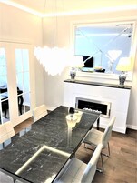 Uma sala de jantar moderna com mesa de mármore, cadeiras brancas, lareira e uma grande janela com vista.