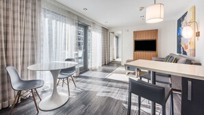 Quarto de hotel moderno com área de refeições, sofá, televisão na parede e janelas amplas com cortinas.