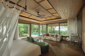 Quarto espaçoso com uma cama grande, um banquinho verde, uma mesinha e vista para uma piscina e vegetação exuberante.