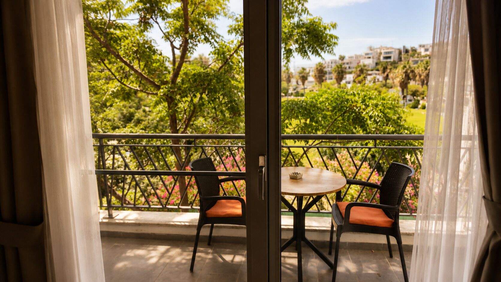 Uma varanda com mesa e cadeiras, vista para áreas verdes e edifícios, e uma porta com cortinas.
