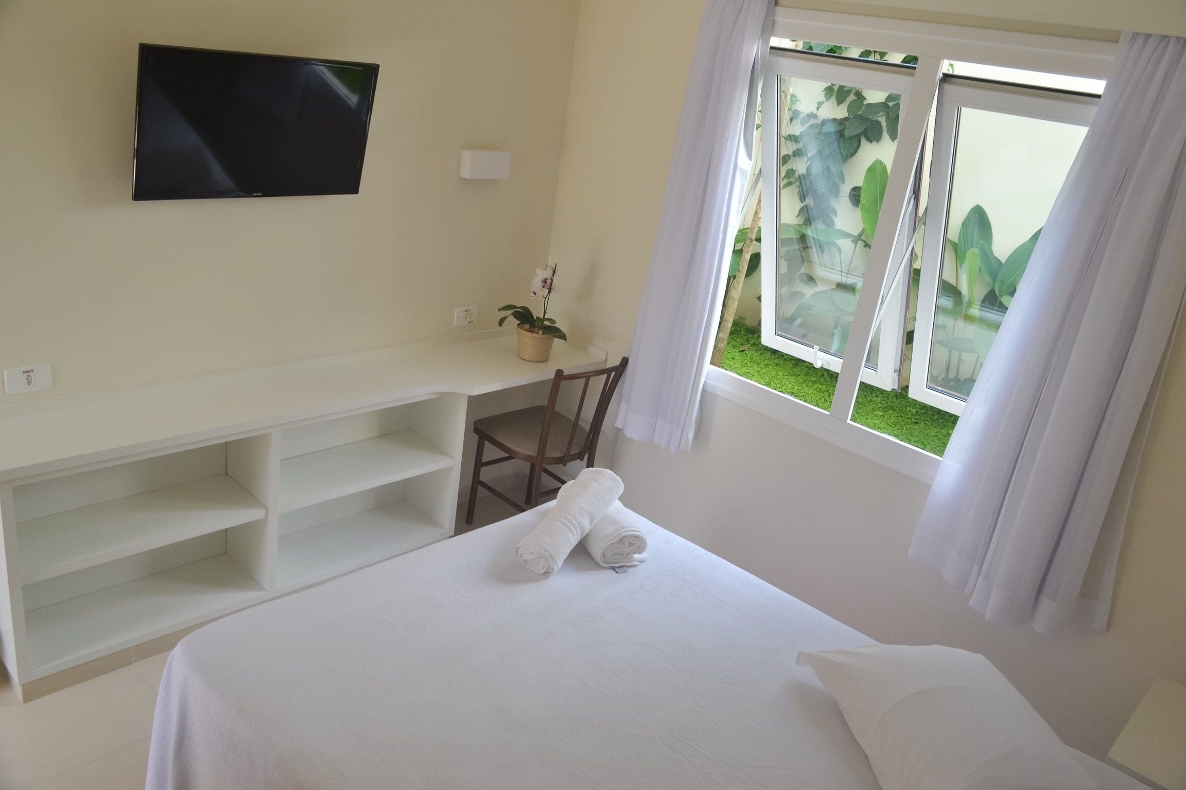 Quarto de hotel com cama, escrivaninha com cadeira, televisão na parede e janela com cortinas.