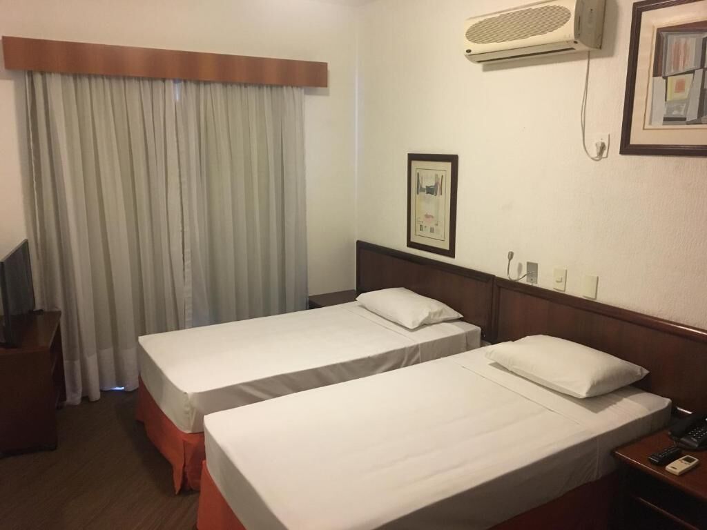 Quarto de hotel com duas camas de solteiro, cabeceira de madeira, mesa de cabeceira com telefone, quadro na parede e aparelho de ar condicionado.