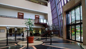 Um hall amplo com teto alto, um vaso de planta no centro, uma escadaria e uma grande janela com vista para o exterior.