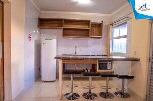 Uma cozinha compacta com geladeira, micro-ondas e armários de madeira. Há três banquetas e uma janela que oferece luz natural.
