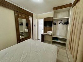 Quarto de hotel com cama, guarda-roupa com roupas penduradas, televisão e espelho.