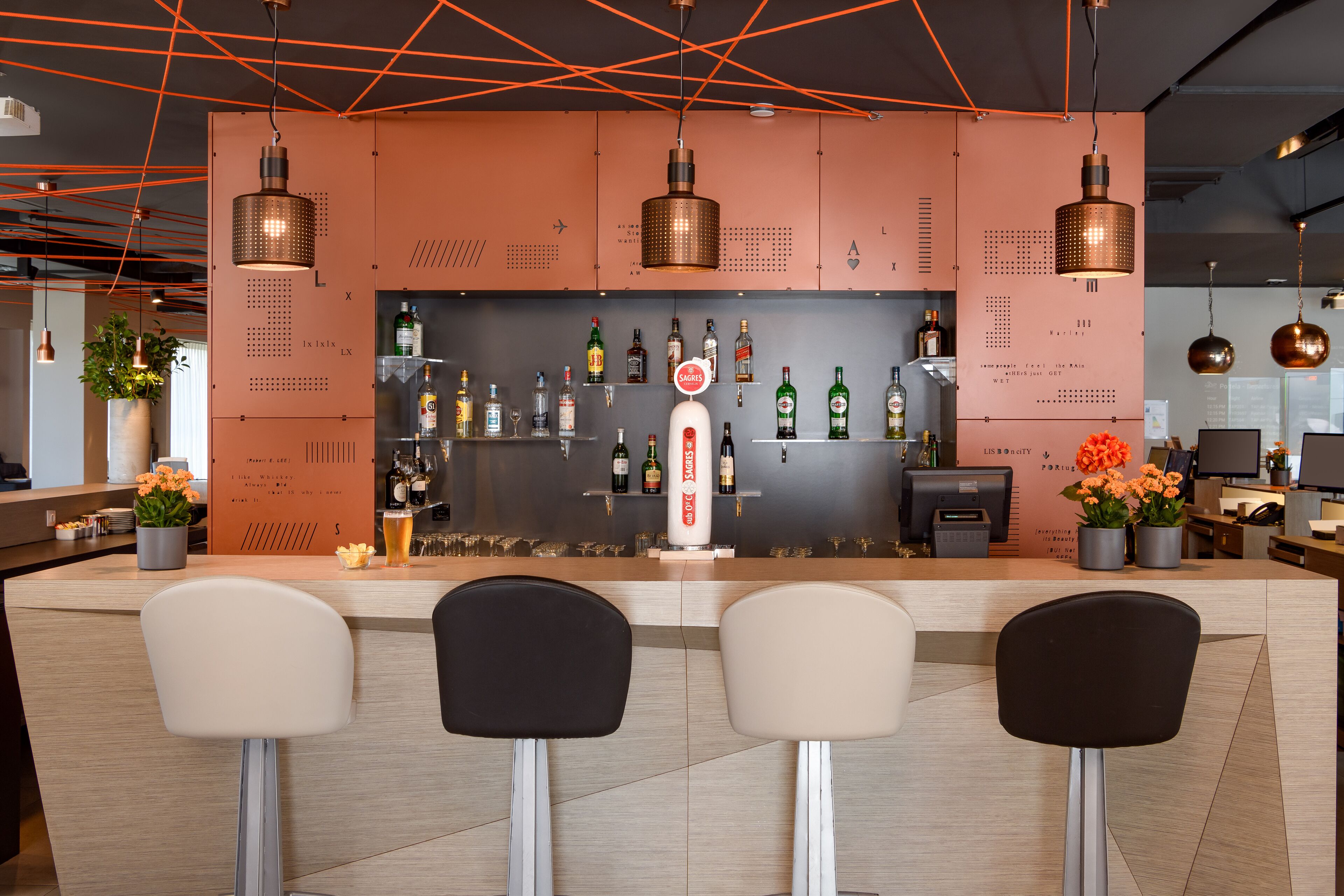 Uma área de bar com três banquetas, variedade de garrafas de bebidas e elementos decorativos como plantas e flores em vasos.