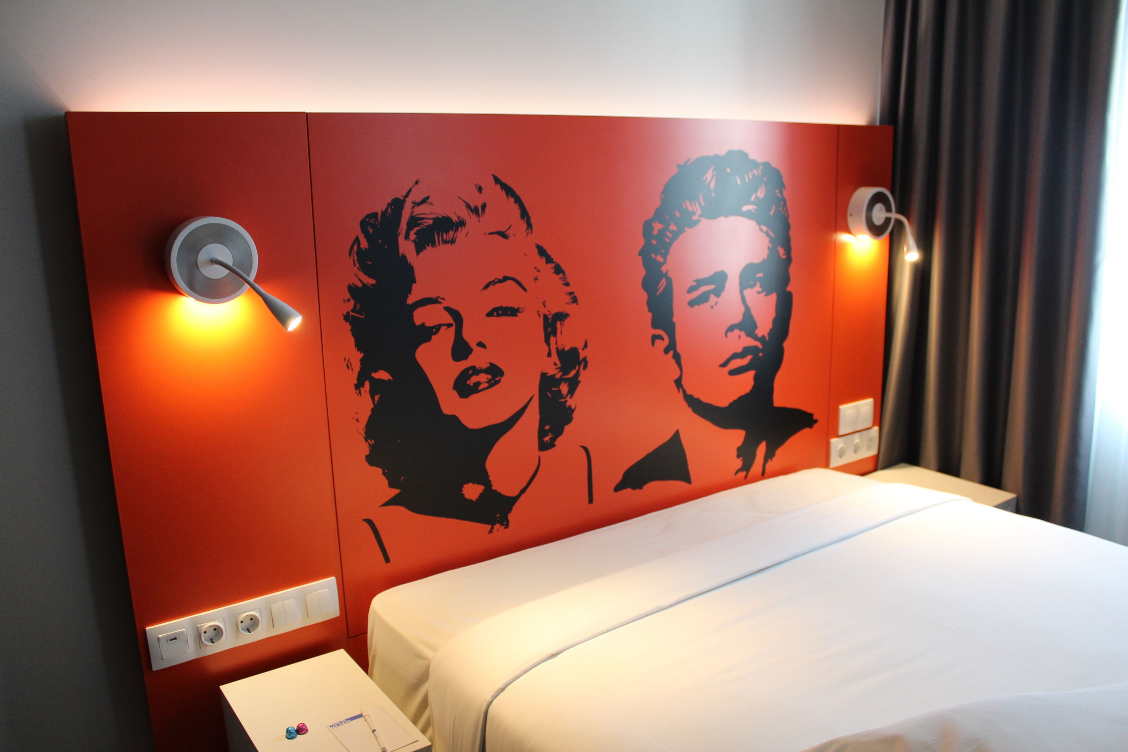 Quarto de hotel com uma cama, luminária na parede e tomada.