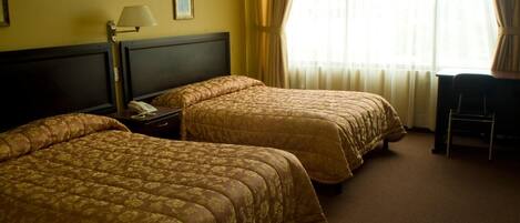 Quarto de hotel com duas camas, uma escrivaninha, uma cadeira e um abajur.