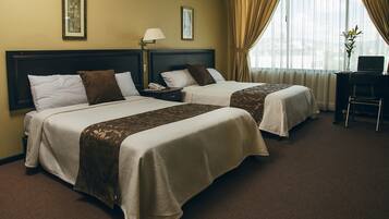 Quarto de hotel com duas camas, uma escrivaninha com cadeira, uma luminária e uma janela com cortinas.