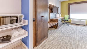 Quarto de hotel com guarda-roupa de madeira, micro-ondas, escrivaninha com cadeira e uma janela grande com persianas.